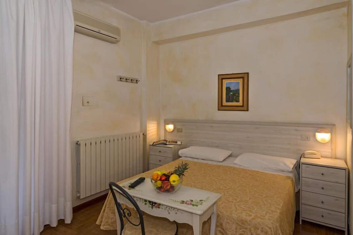 Quarto de hotel com cama, mesas de cabeceira, uma mesinha com uma cesta de frutas, uma cadeira e cortina.