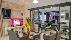 Uma sala de estar moderna com televisão de tela plana, móveis de vime e porta de vidro que dá para um pátio externo com cadeiras e mesa.