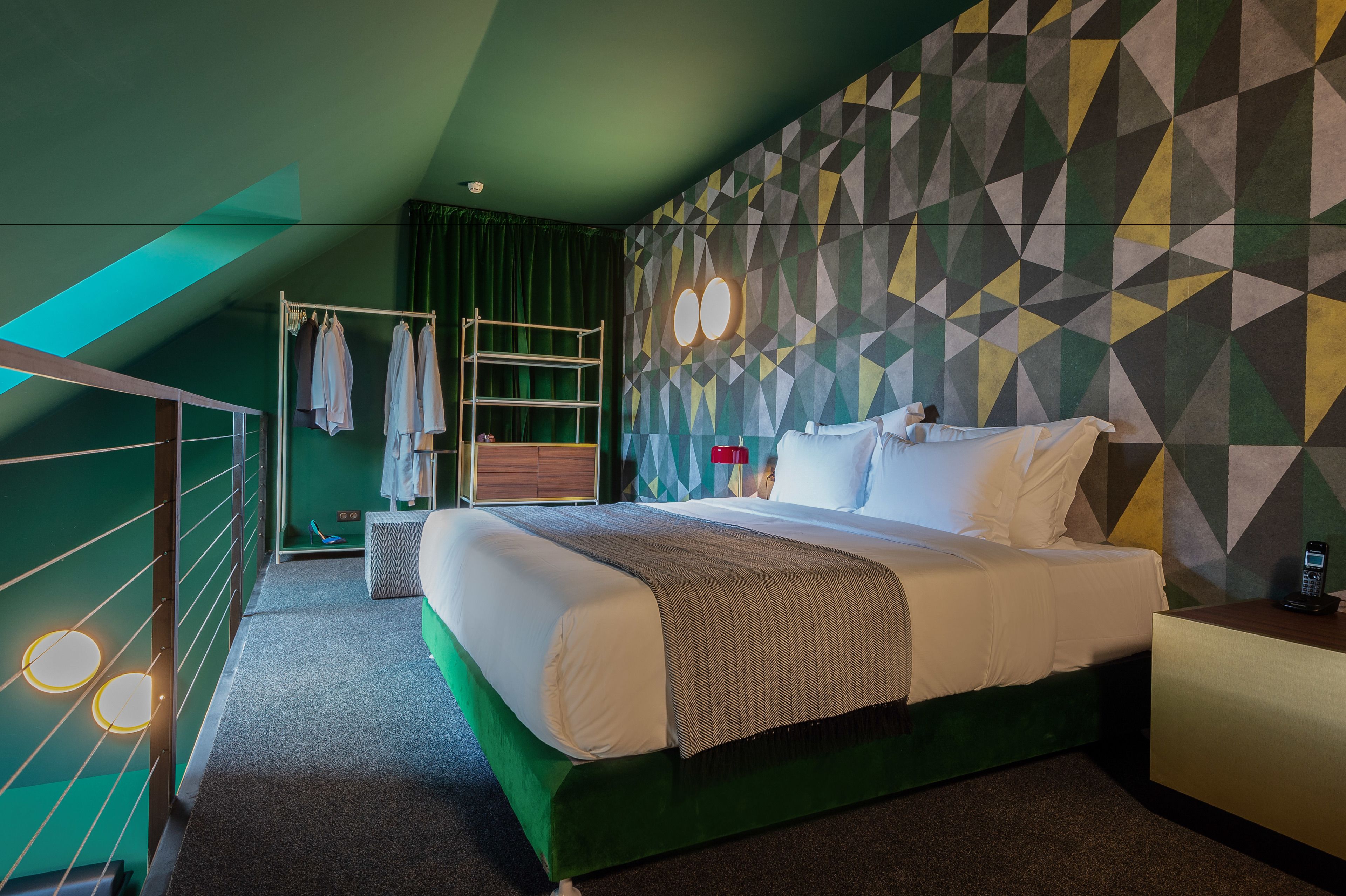 Quarto de hotel moderno com parede de padrão geométrico, cama com roupa de cama branca, mesa de cabeceira com telefone e uma parede verde de destaque.