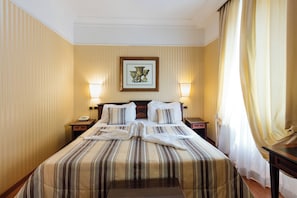 Quarto de hotel com uma cama grande, duas mesas de cabeceira, um criado-mudo, um quadro na parede e uma janela com cortinas.