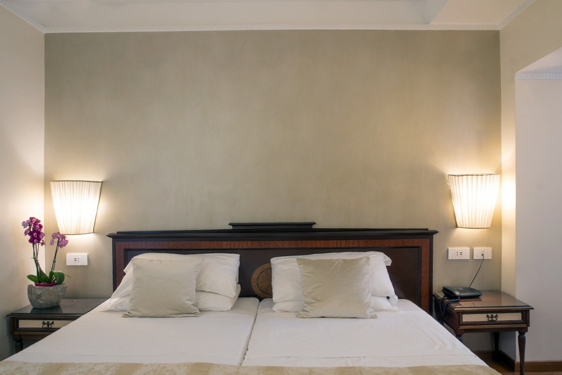 Quarto de hotel com uma cama grande, duas luminárias de cabeceira, mesa de cabeceira com telefone e um vaso de planta.