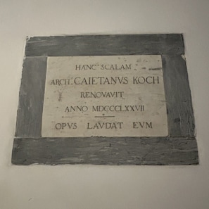 Uma placa com texto, possivelmente histórica ou comemorativa, fixada em uma parede.