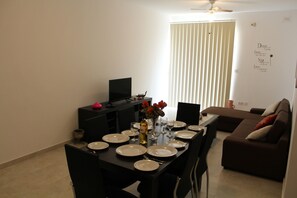 Uma sala de jantar com uma mesa posta para quatro pessoas, uma televisão em um suporte e um sofá ao fundo.