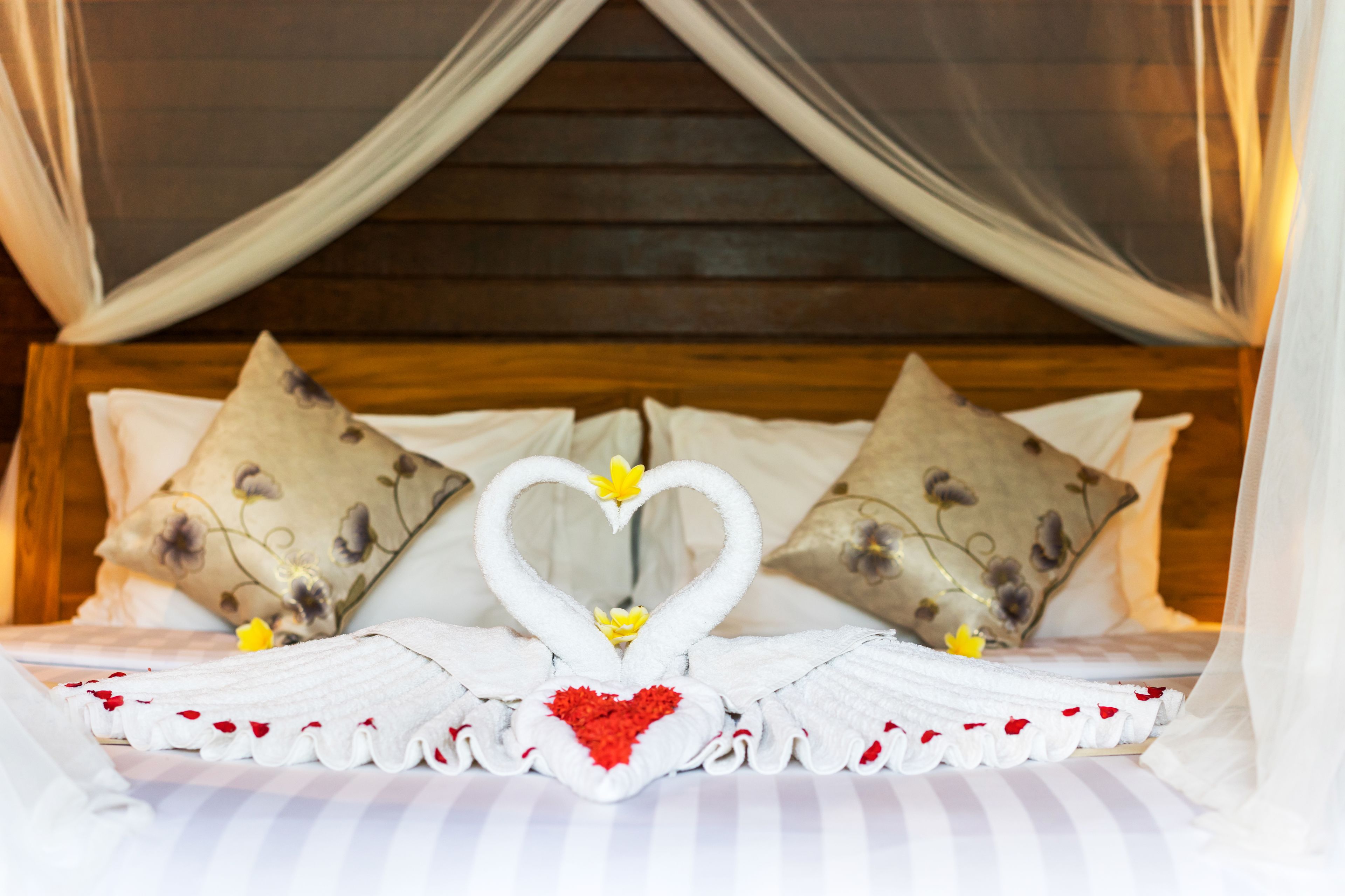 Uma cama com dossel, almofadas brancas com motivos florais e um cisne decorativo feito de toalhas.