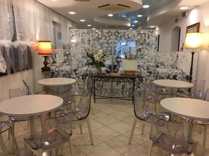 Uma sala de jantar com mesas redondas e cadeiras transparentes, uma parede decorativa com motivos florais e uma mesa central com um vaso de flores.