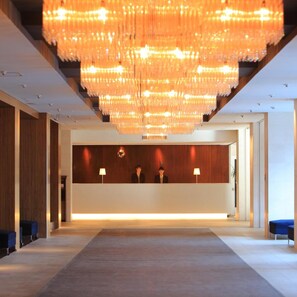 Uma área de recepção de hotel com um grande candelabro, um balcão de recepção e duas luminárias de cada lado.