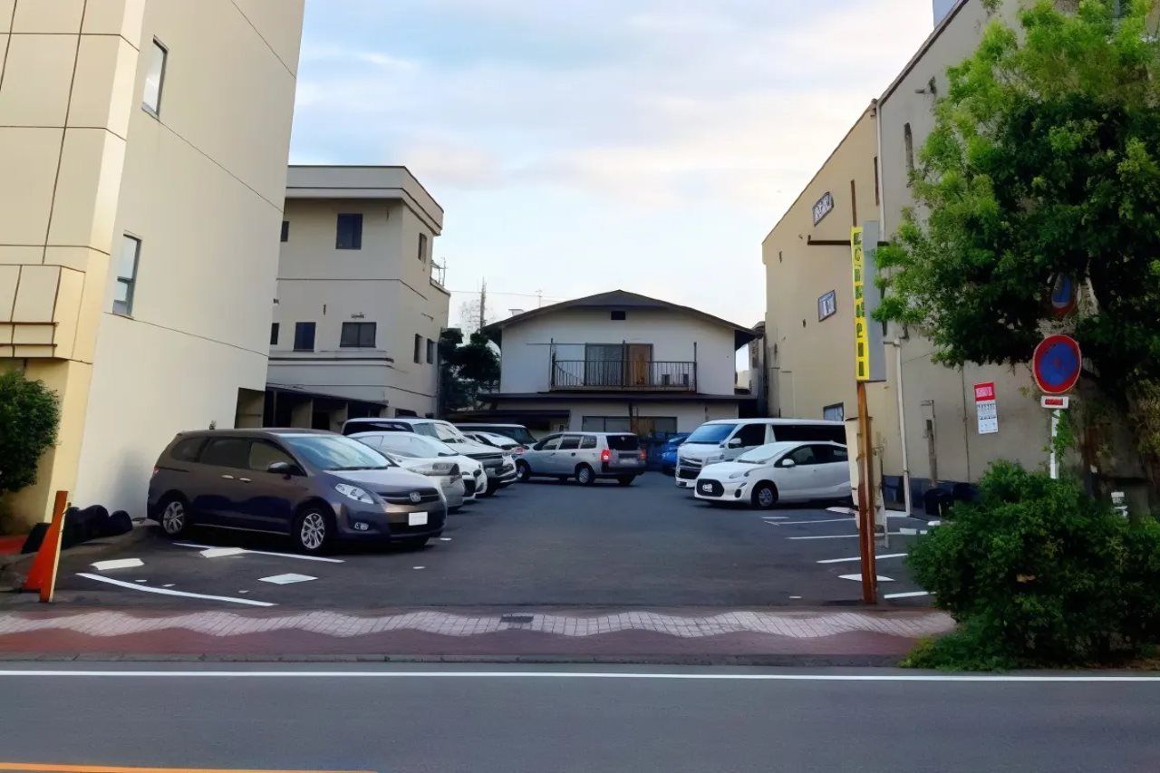 Um estacionamento com vários carros estacionados em fileiras, um edifício com telhado plano e varanda, e céu claro.