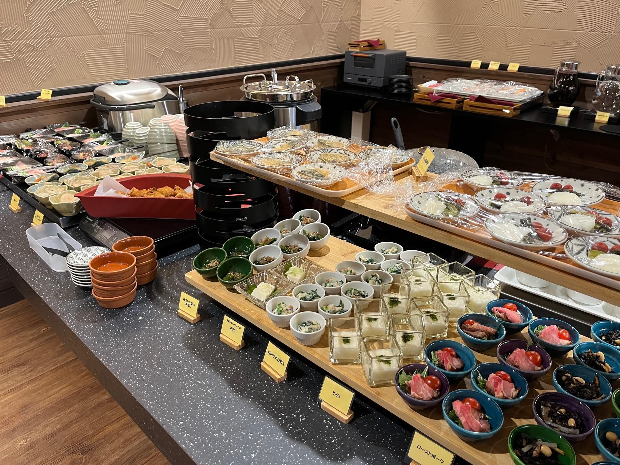 Buffet variado com diversos pratos e bebidas, incluindo recipientes com comida, pratos e uma máquina de café.
