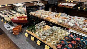 Buffet variado com diversos pratos e bebidas, incluindo recipientes com comida, pratos e uma máquina de café.