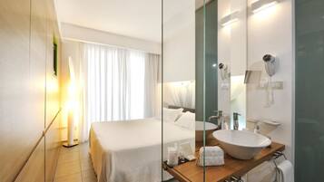 Quarto de hotel com uma cama grande, uma mesinha de cabeceira de madeira com artigos de higiene pessoal, um box de chuveiro envidraçado e um espelho grande.