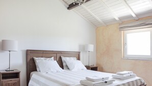 Uma cama bem arrumada com lençóis brancos, duas mesas de cabeceira com abajures e uma janela com vista para o exterior.