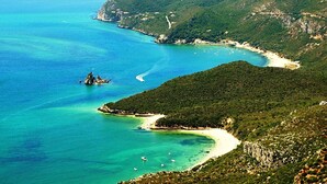 Paisagem costeira com vista clara do mar, uma estrada sinuosa e uma pequena ilha com afloramentos rochosos.