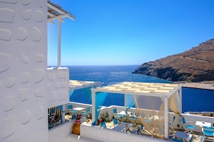 Terraço com paredes brancas e detalhes em azul, com vista para o mar azul e um litoral rochoso.