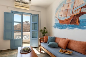 Quarto com sofá de madeira, mesa com uma planta e um prato de doces, um mural de um navio a vela, e vista através de uma porta para uma varanda com um edifício branco.