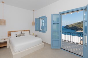 Quarto com cama, mesa de cabeceira e vista para o mar através de uma porta aberta.