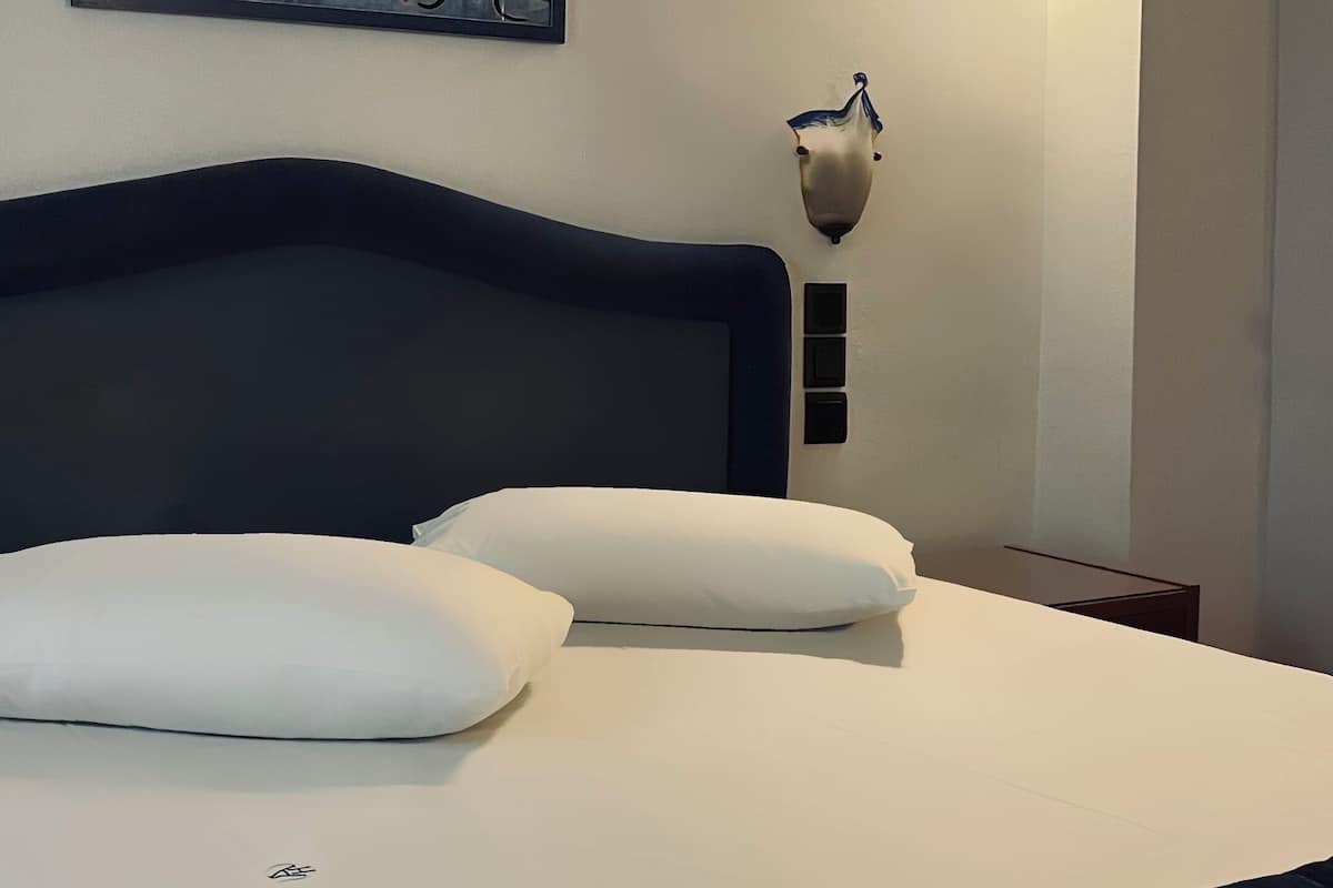Cama bem arrumada com lençóis brancos e cabeceira escura, obra de arte abstrata na parede e luminária fixada na parede.