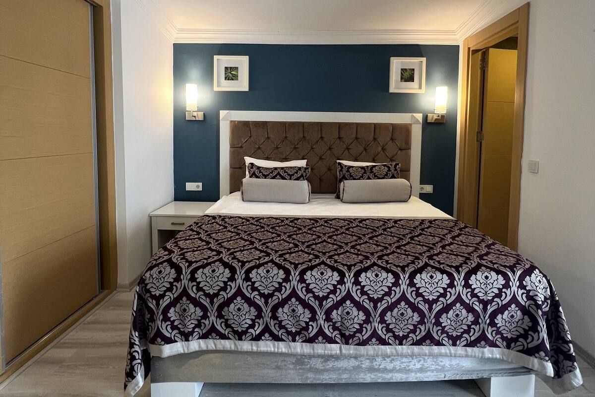 Quarto com cabeceira estofada, colcha estampada, duas mesas de cabeceira com abajures e piso de madeira.