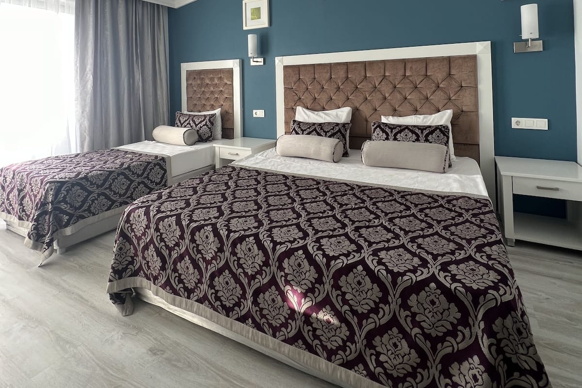 Quarto de hotel com duas camas, uma parede azul de destaque e uma janela com cortinas.