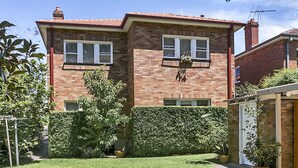 Uma casa de dois andares de tijolo com telhado vermelho, venezianas brancas e um pequeno jardim com sebes e uma árvore.