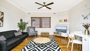 Uma sala de estar com um sofá preto, uma poltrona cinza, uma mesa de centro redonda de madeira, uma TV e um ventilador de teto.