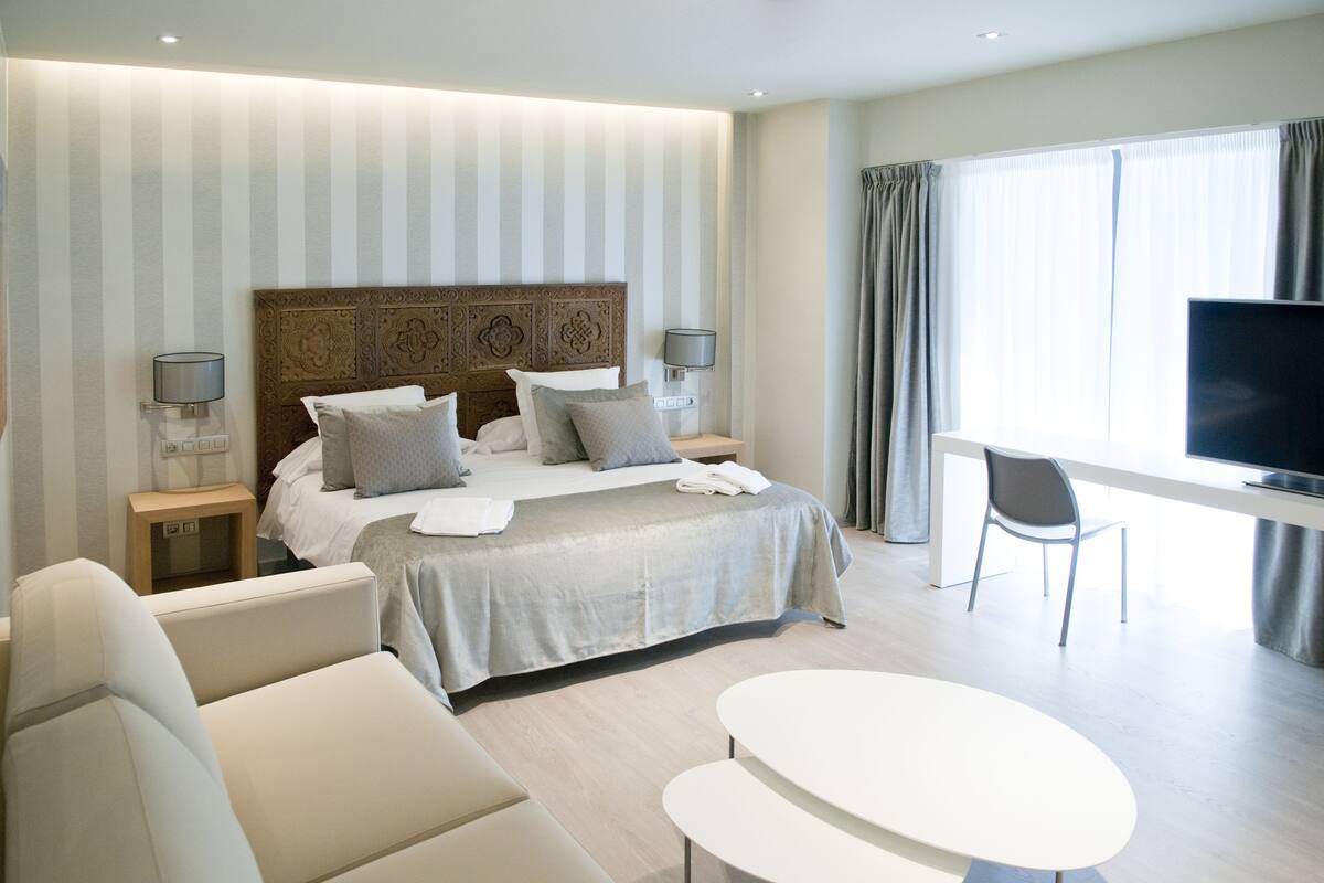 Quarto de hotel moderno com uma cama grande, duas mesas de cabeceira com abajures, uma TV, um espaço de estar com sofá e uma mesa pequena, e uma janela ampla com cortinas.