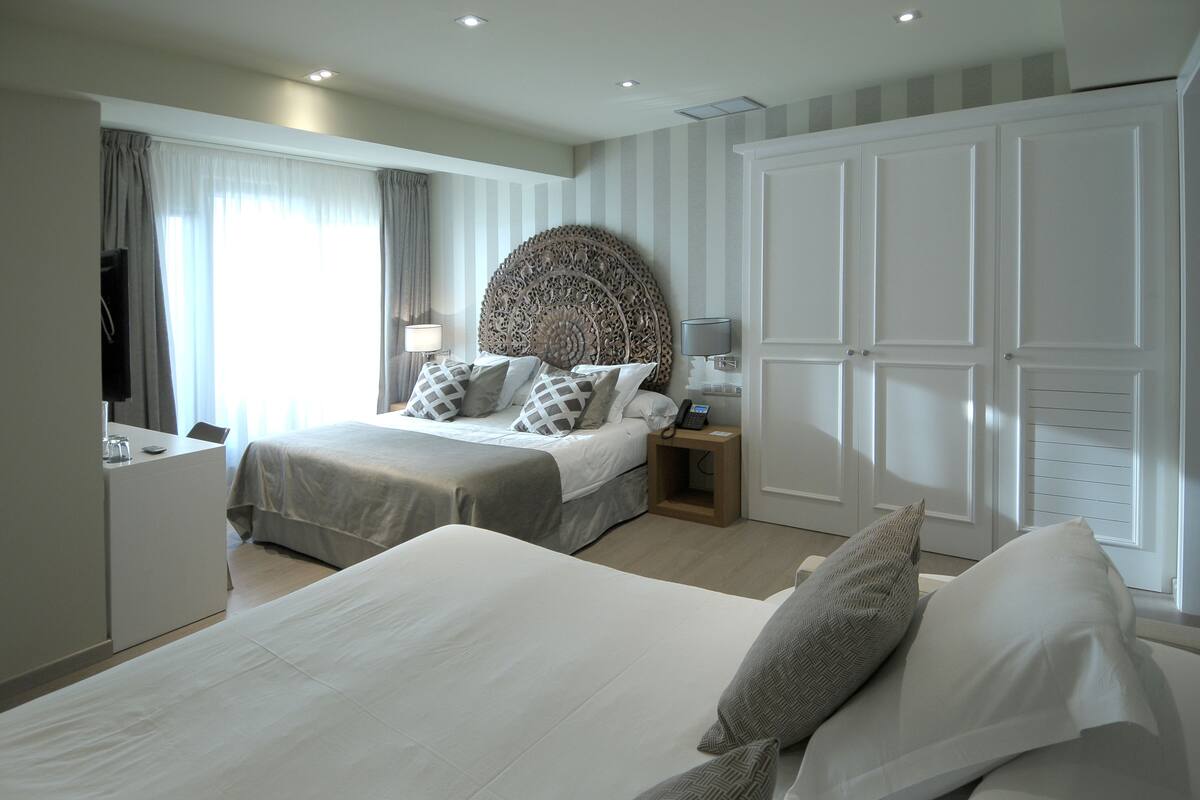 Quarto de hotel com duas camas, uma cabeceira grande e decorativa, um guarda-roupa e uma escrivaninha com luminária.