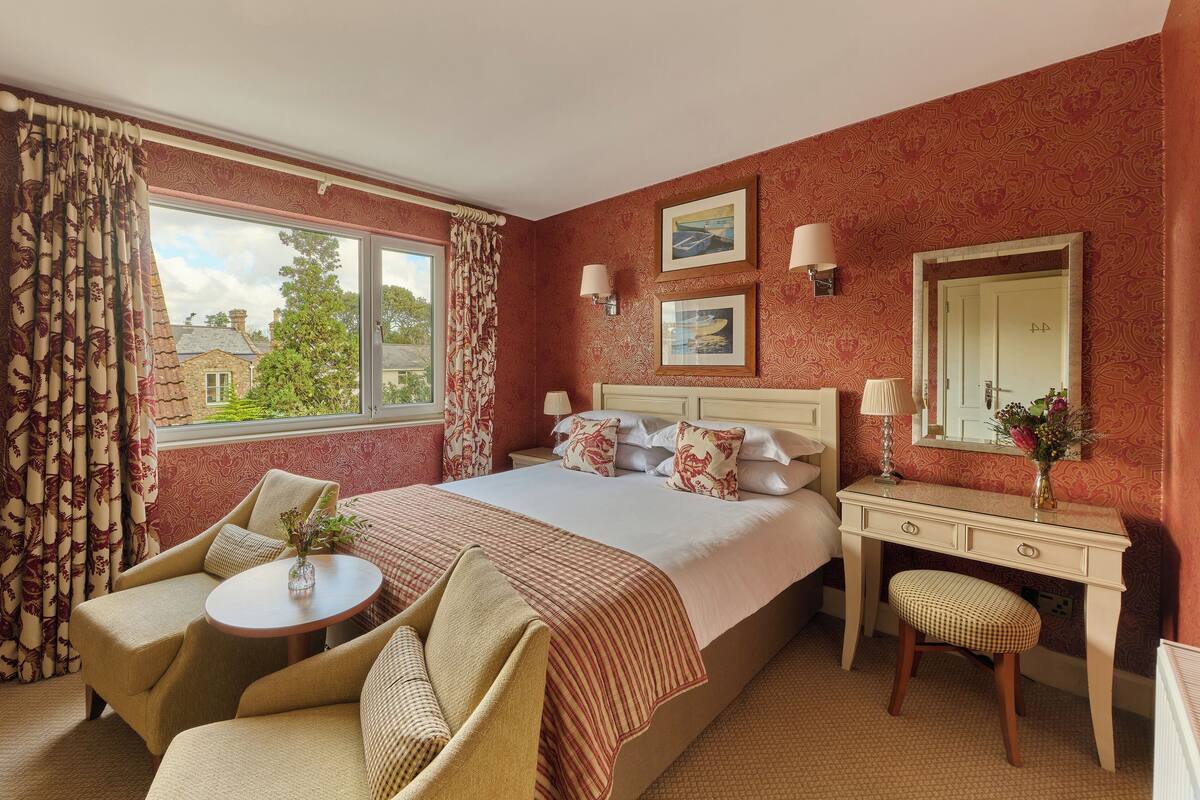 Quarto de hotel com uma cama grande, área de estar com mesa, pequena escrivaninha e cadeira. Há uma janela com cortinas, um espelho e um quadro na parede.