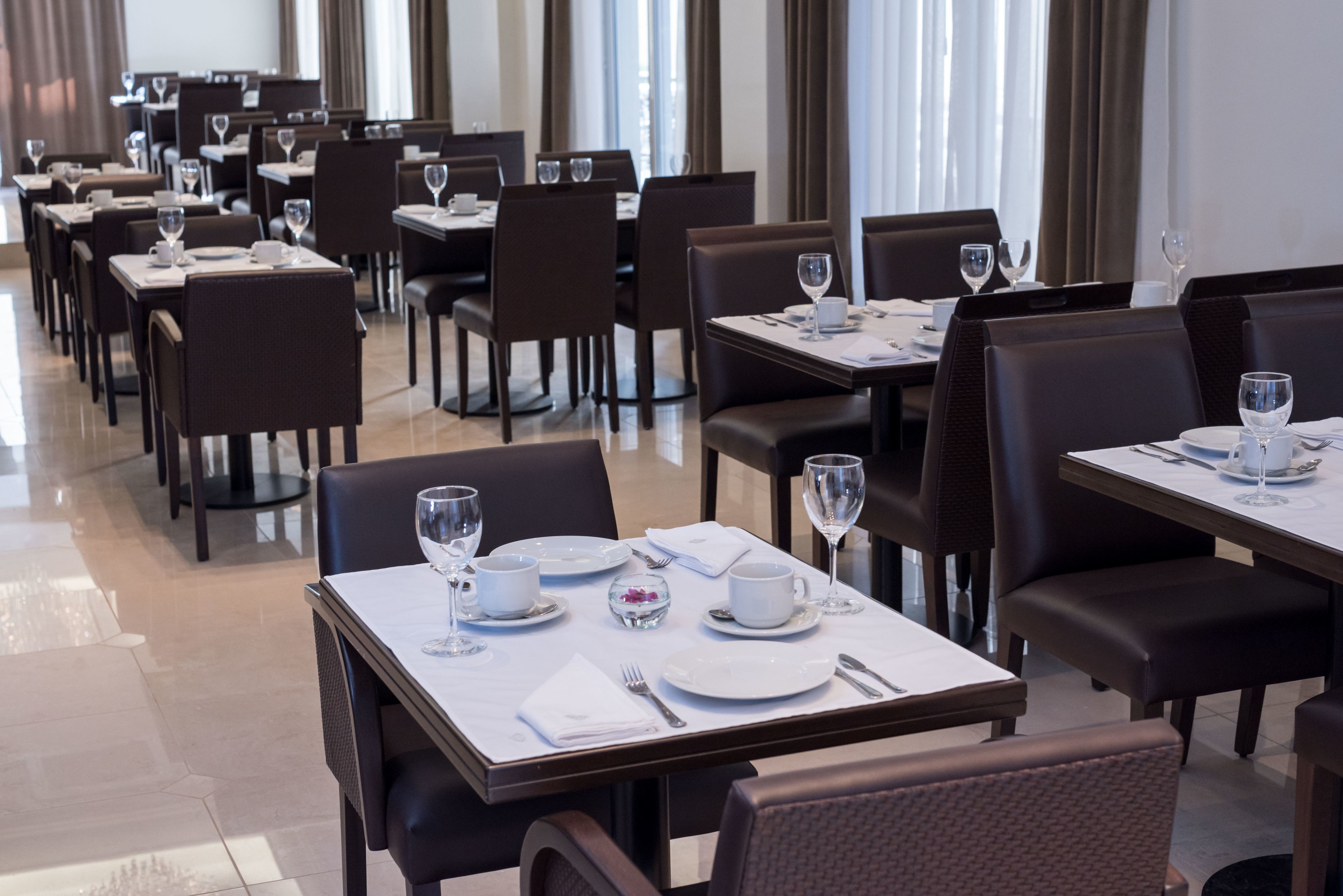 Uma área de refeições de hotel com várias mesas preparadas para o jantar, cada uma com cadeiras, pratos, talheres e copos de vinho.
