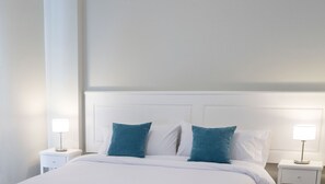 Cama bem arrumada com roupa de cama branca e duas almofadas azul-turquesa, acompanhada por duas mesas de cabeceira com abajures.