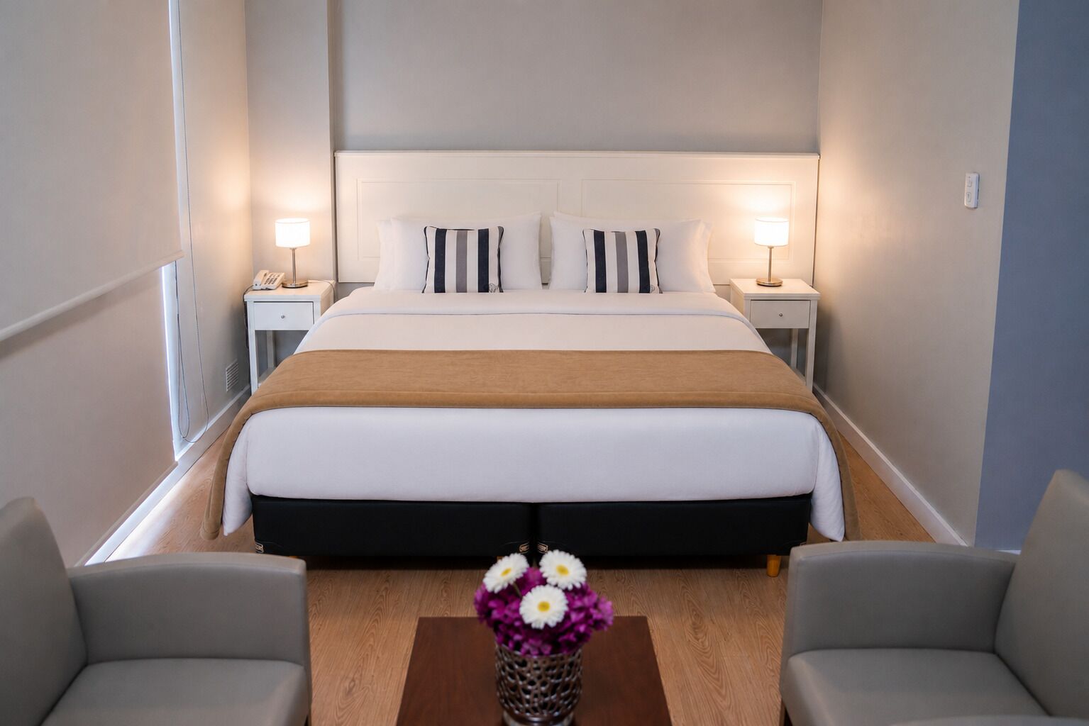 Quarto de hotel com uma cama grande, luminárias de cabeceira, um vaso com flores e duas poltronas.