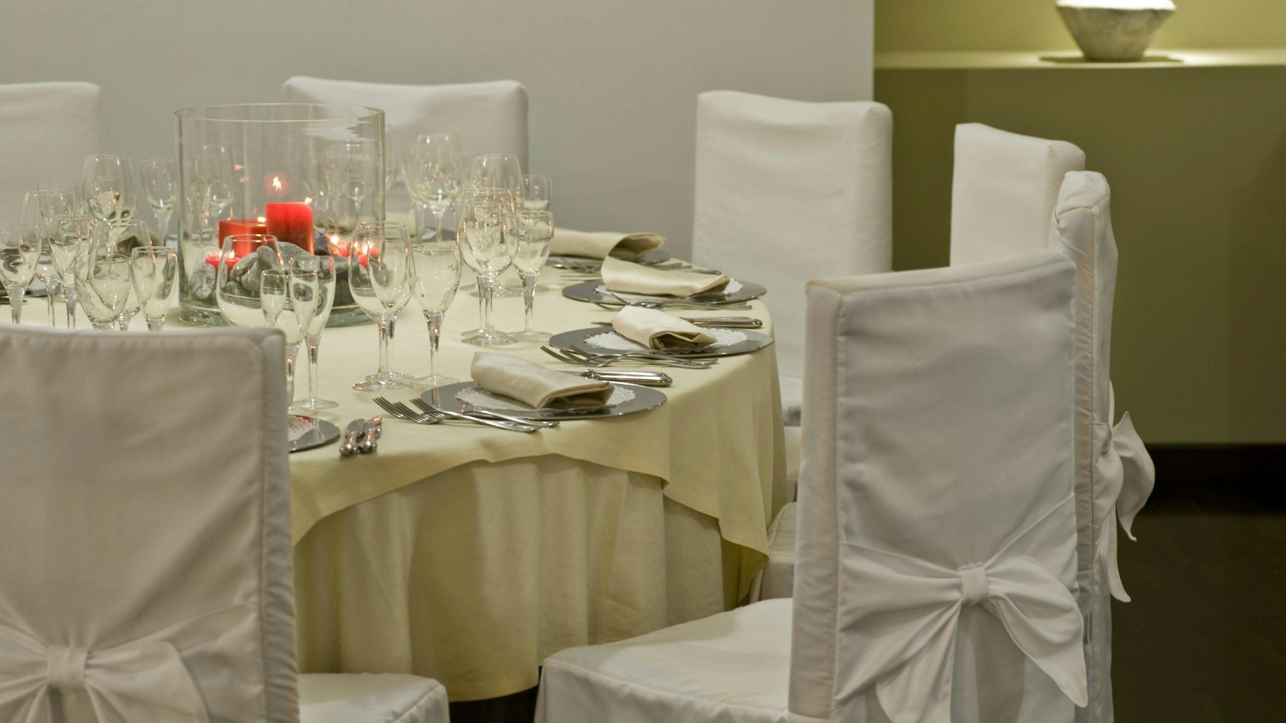 Mesa de jantar formal posta com toalha branca, copos e talheres, rodeada por cadeiras brancas decoradas com laços.