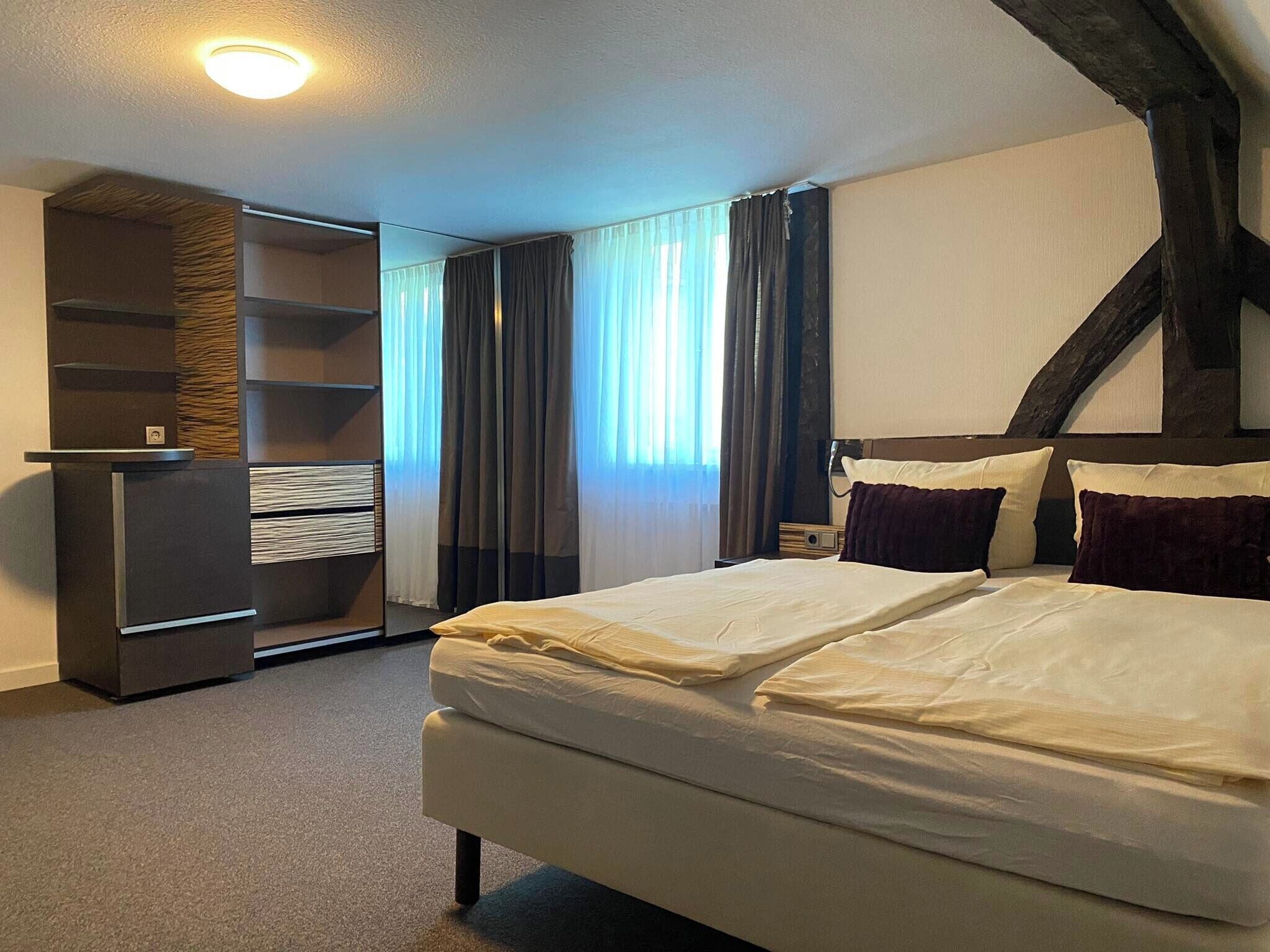 Quarto de hotel com cama, guarda-roupa, espelho e janela com cortinas.