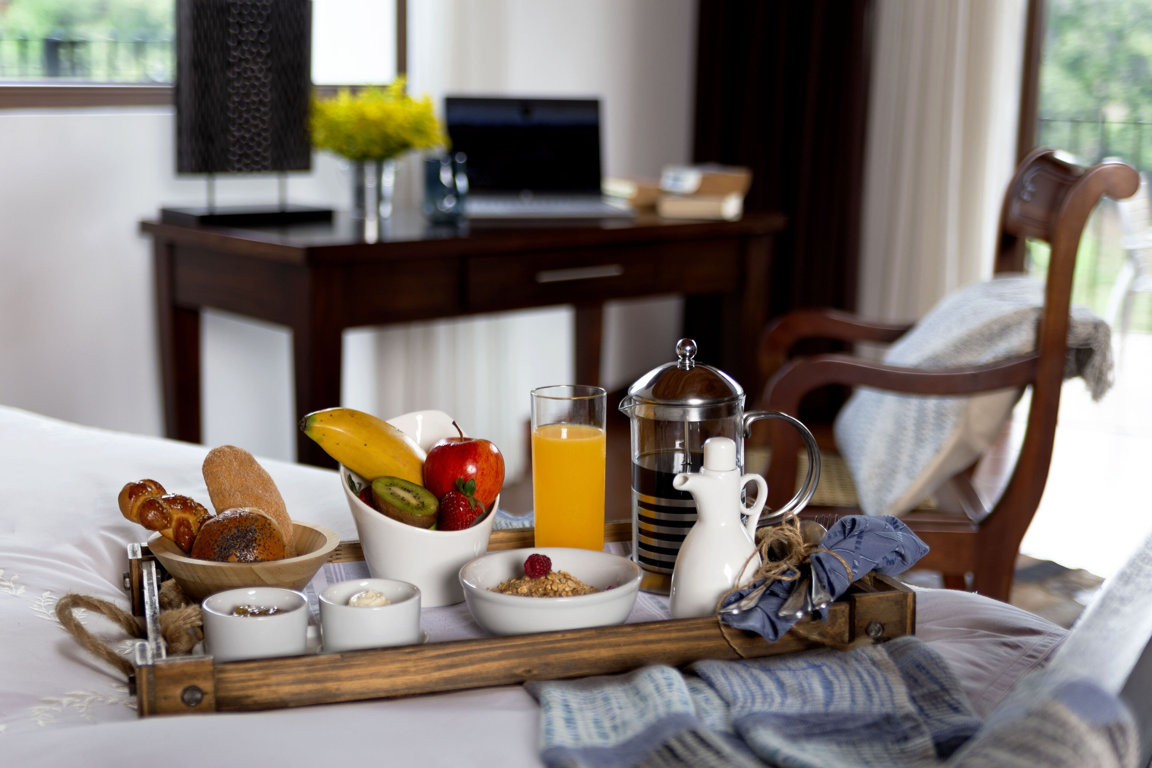 Bandeja de café da manhã com pão, frutas e uma cafeteira em uma cama, com um laptop e uma mesa de trabalho ao fundo.