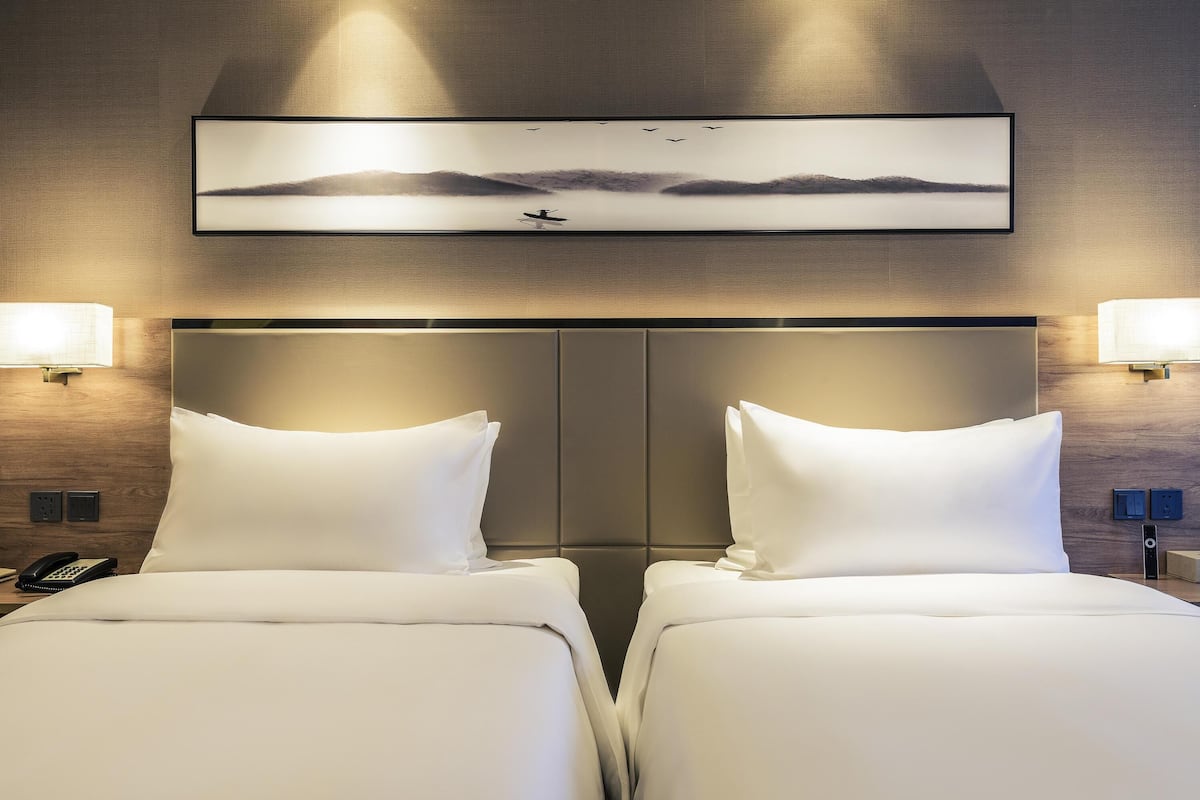 Quarto de hotel com duas camas, uma cabeceira grande, luminárias fixadas na parede e um quadro emoldurado sobre as camas.