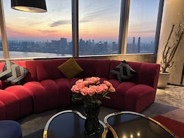 Um sofá vermelho com almofadas estampadas, um vaso com flores cor-de-rosa e vista para o horizonte urbano através de grandes janelas.