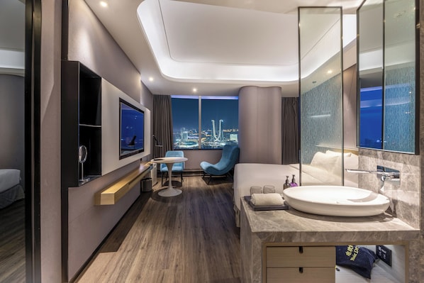 Quarto de hotel moderno com uma cama grande, pia de banheiro redonda, televisão, escrivaninha e vista para a cidade.