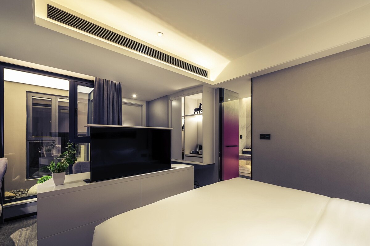Quarto de hotel moderno com cama, televisão de tela plana, escrivaninha e banheiro visível por meio de um painel de vidro.