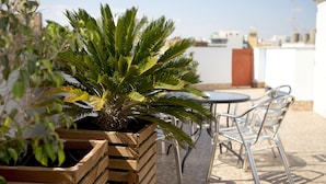 Um terraço no telhado com um cachepot de madeira contendo uma palmeira, cadeiras de metal e uma mesa.