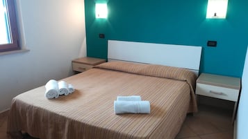 Uma cama bem arrumada com roupa de cama listrada e duas toalhas enroladas, uma mesa de cabeceira com gaveta, e um aplique de parede.