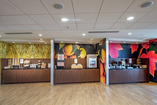 Uma área de recepção moderna com balcões de madeira, um mural com tema de frutas e um espaço de bebidas de autoatendimento.