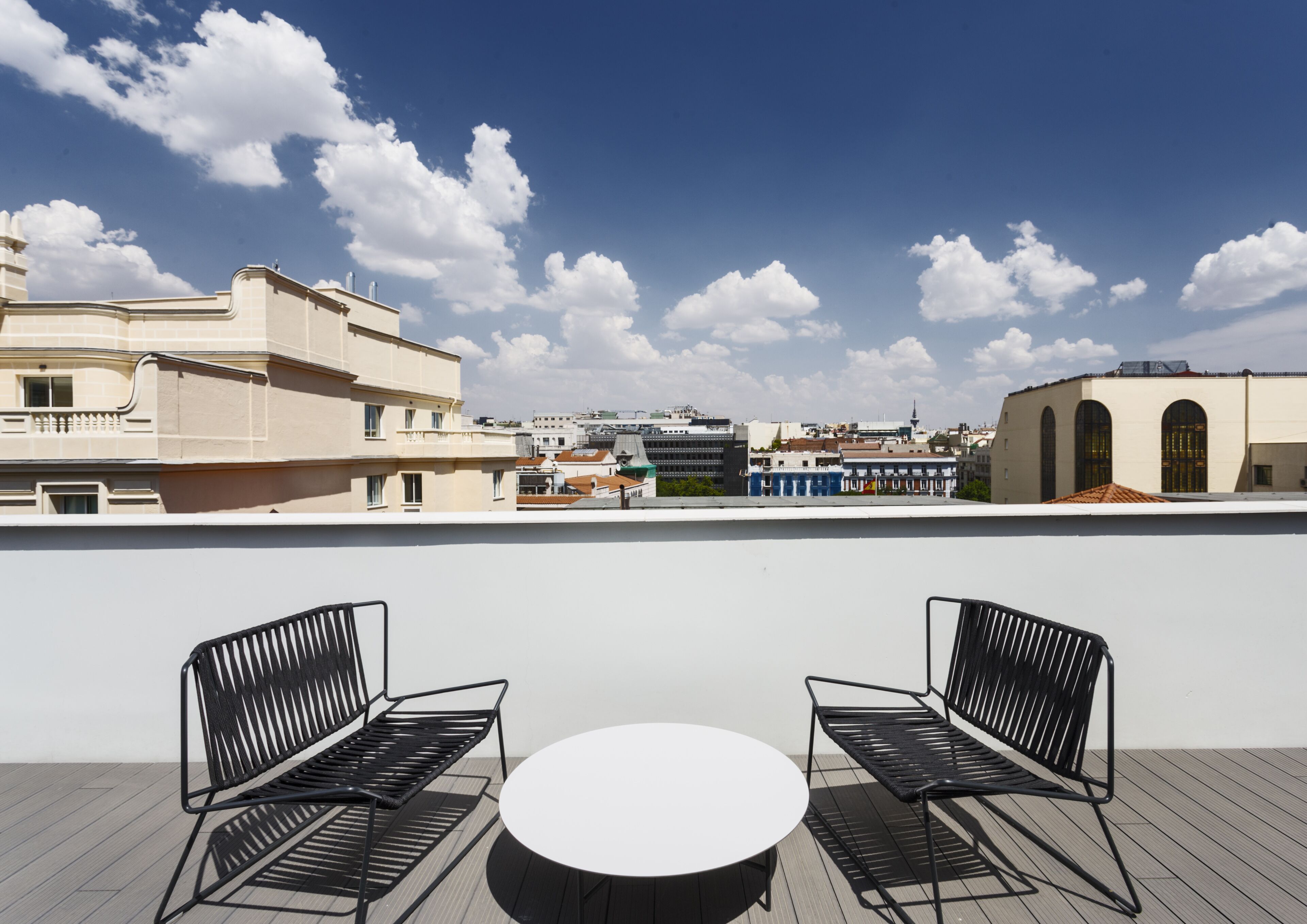 Terraço no telhado com duas cadeiras de metal preto e uma mesa redonda, com vista para uma paisagem urbana com edifícios e céu claro.