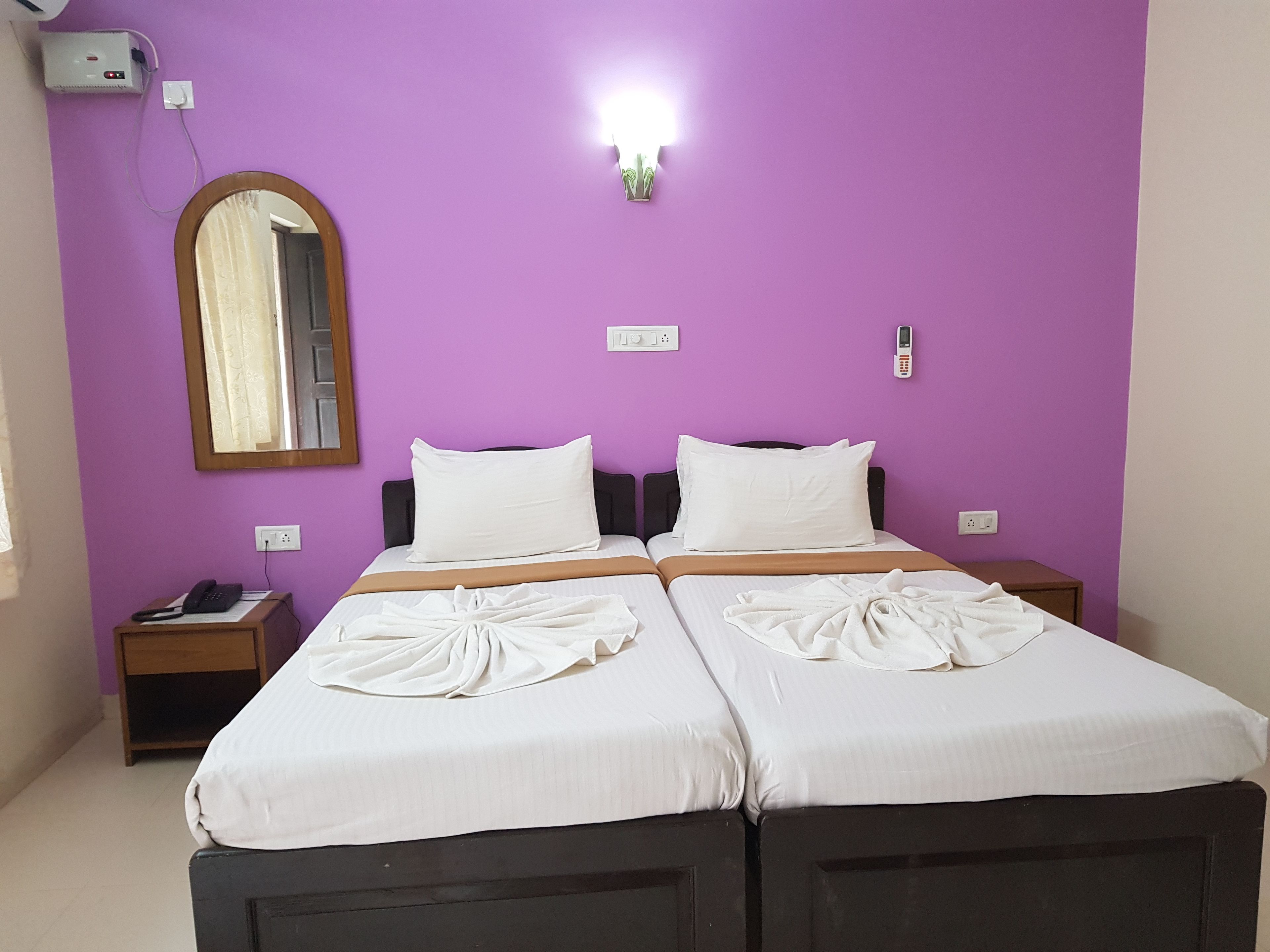 Quarto com paredes roxas, duas camas com lençóis brancos, cabeceira de madeira, mesa de cabeceira com telefone, luminária fixa na parede e um espelho.