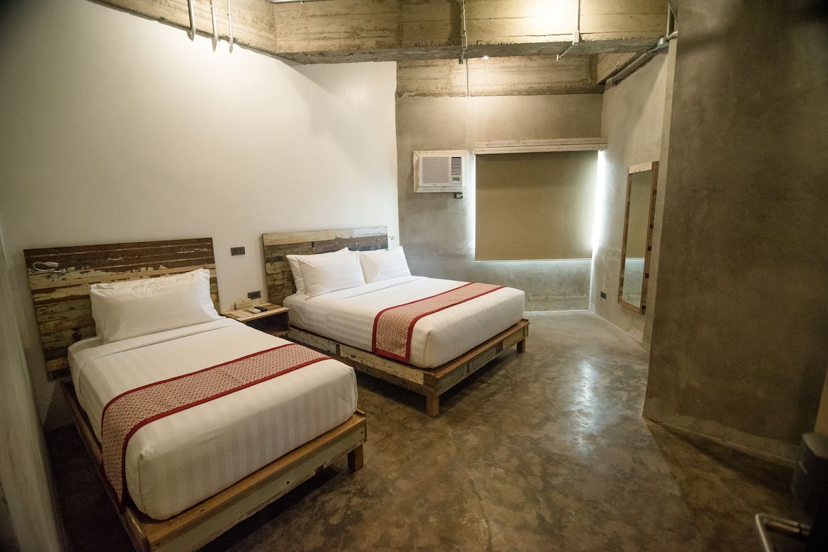 Quarto com duas camas, cada uma com estrutura de madeira e roupa de cama branca. Pequena mesa e cadeira ao lado de cada cama. O chão é de concreto e o design é simples e funcional.