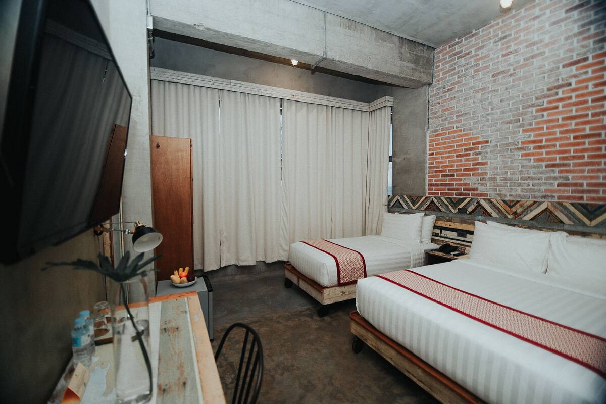 Quarto de hotel com duas camas, uma escrivaninha com cadeira, um espelho e parede com padrão de tijolos.