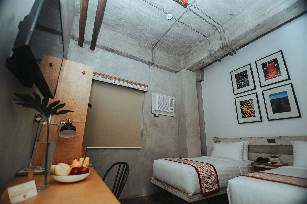 Quarto com duas camas, uma mesa de madeira com uma tigela de frutas, um espelho e um respiradouro fixado na parede.