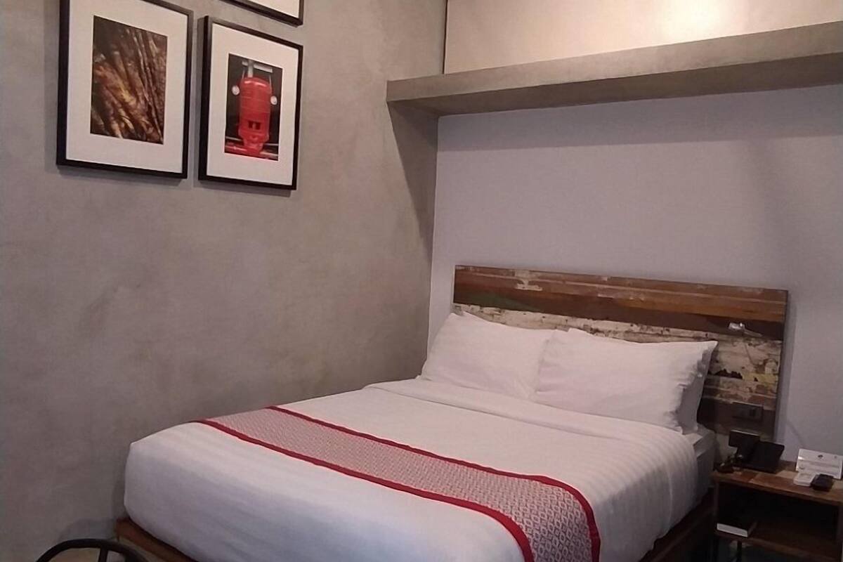 Uma cama de solteiro com cabeceira de madeira, uma colcha vermelha e branca e uma pequena mesa de cabeceira com uma luminária. Quatro quadros emoldurados estão pendurados na parede acima da cama.