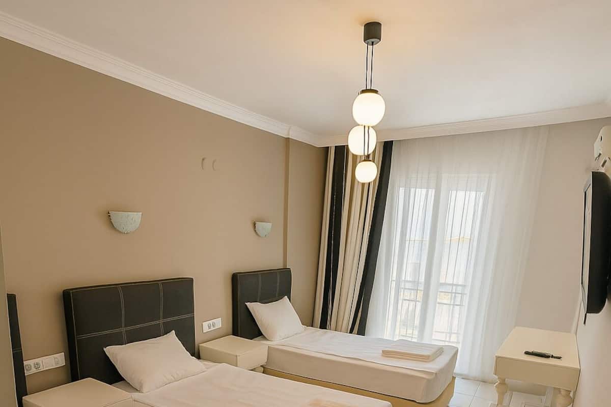 Quarto de hotel com três camas, uma pequena escrivaninha, um espelho e um lustre.