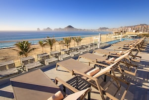 Uma área de estar no terraço com espreguiçadeiras e piscina, com vista para uma praia e montanhas.