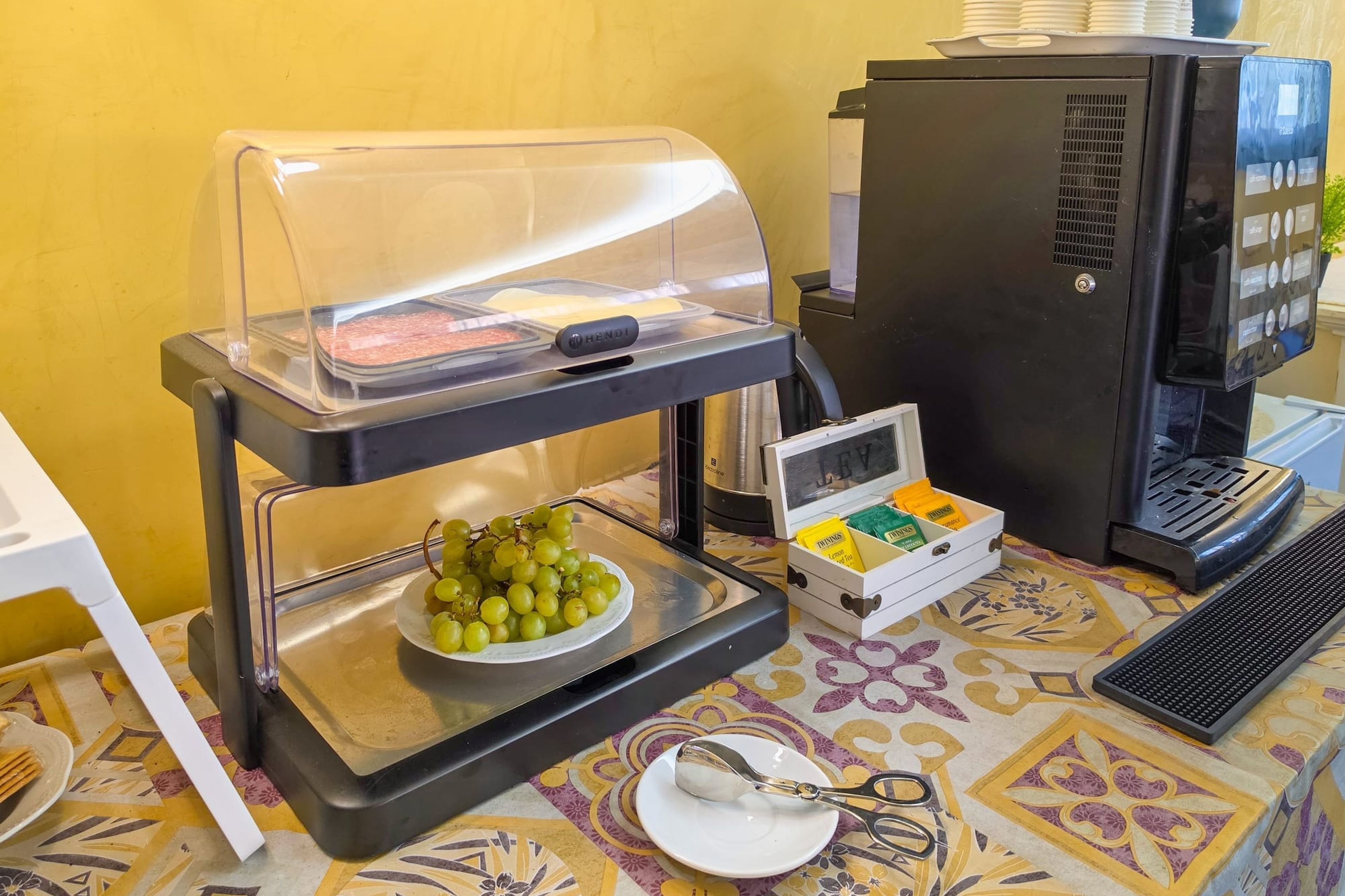 Disposição de buffet com um display de comida coberto, uma máquina de café e uma bandeja de uvas.