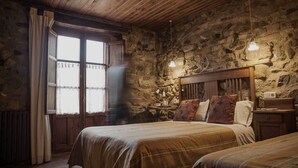 Quarto rústico com parede de pedra, teto de madeira e cama com cabeceira de madeira. Há uma janela com cortinas, uma mesa com abajur e uma mesa de cabeceira com abajur.